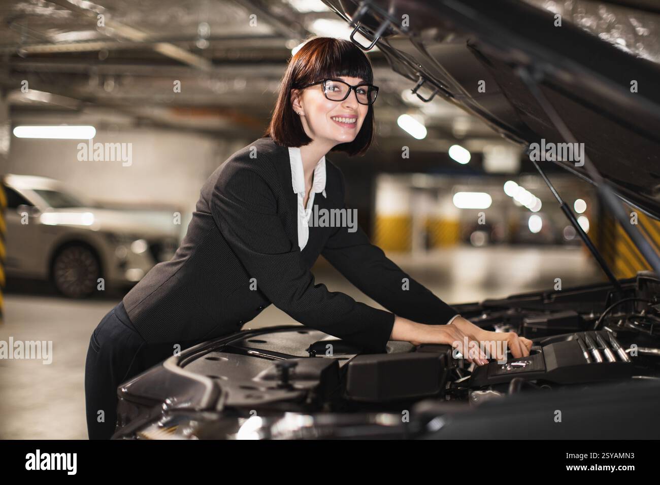 Confident Caucasian businesswoman in her 30s inspects car engine under hood in underground ...