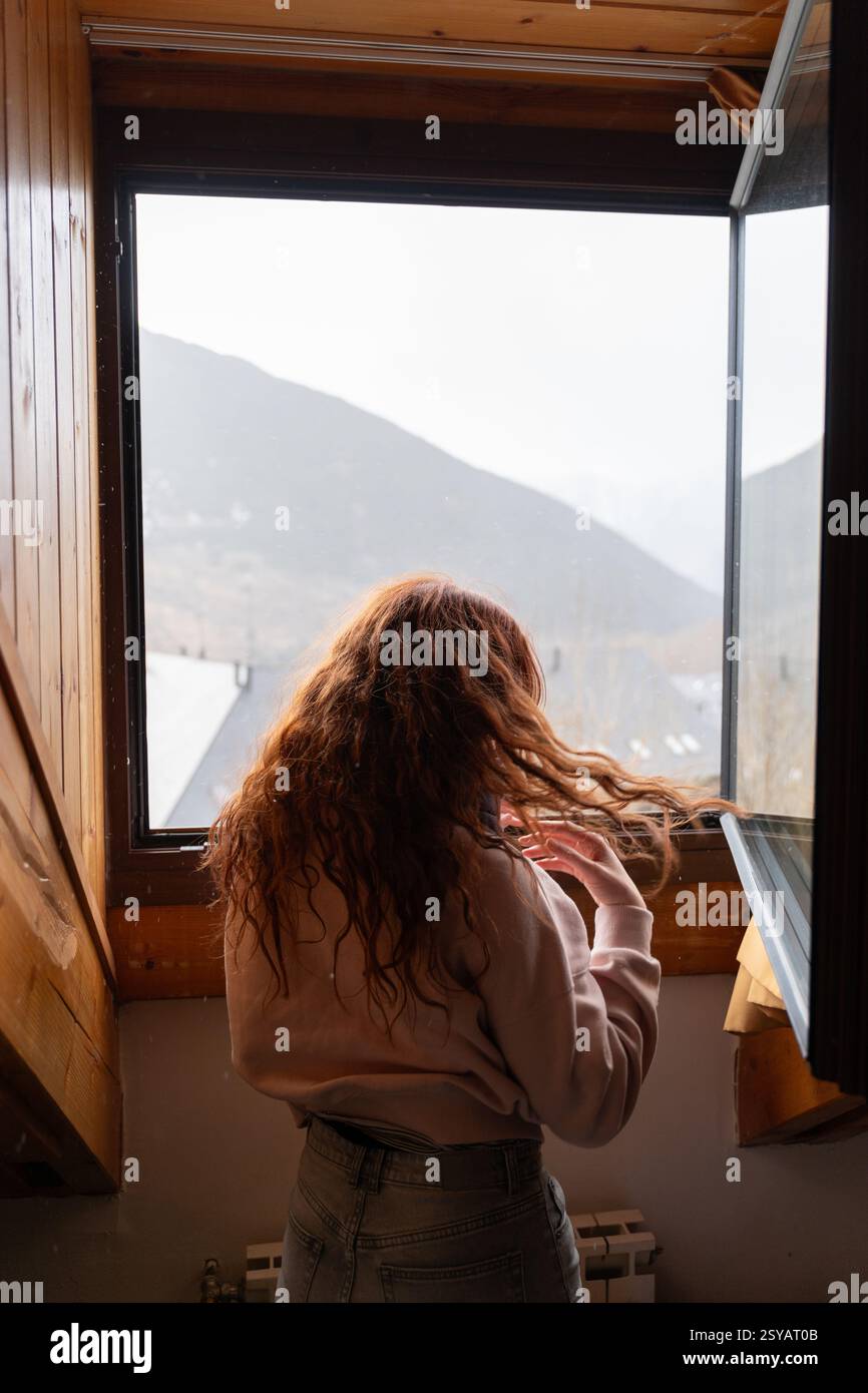 Back view of an unrecognizable woman with red hair standing by a window, gazing at mountain ...