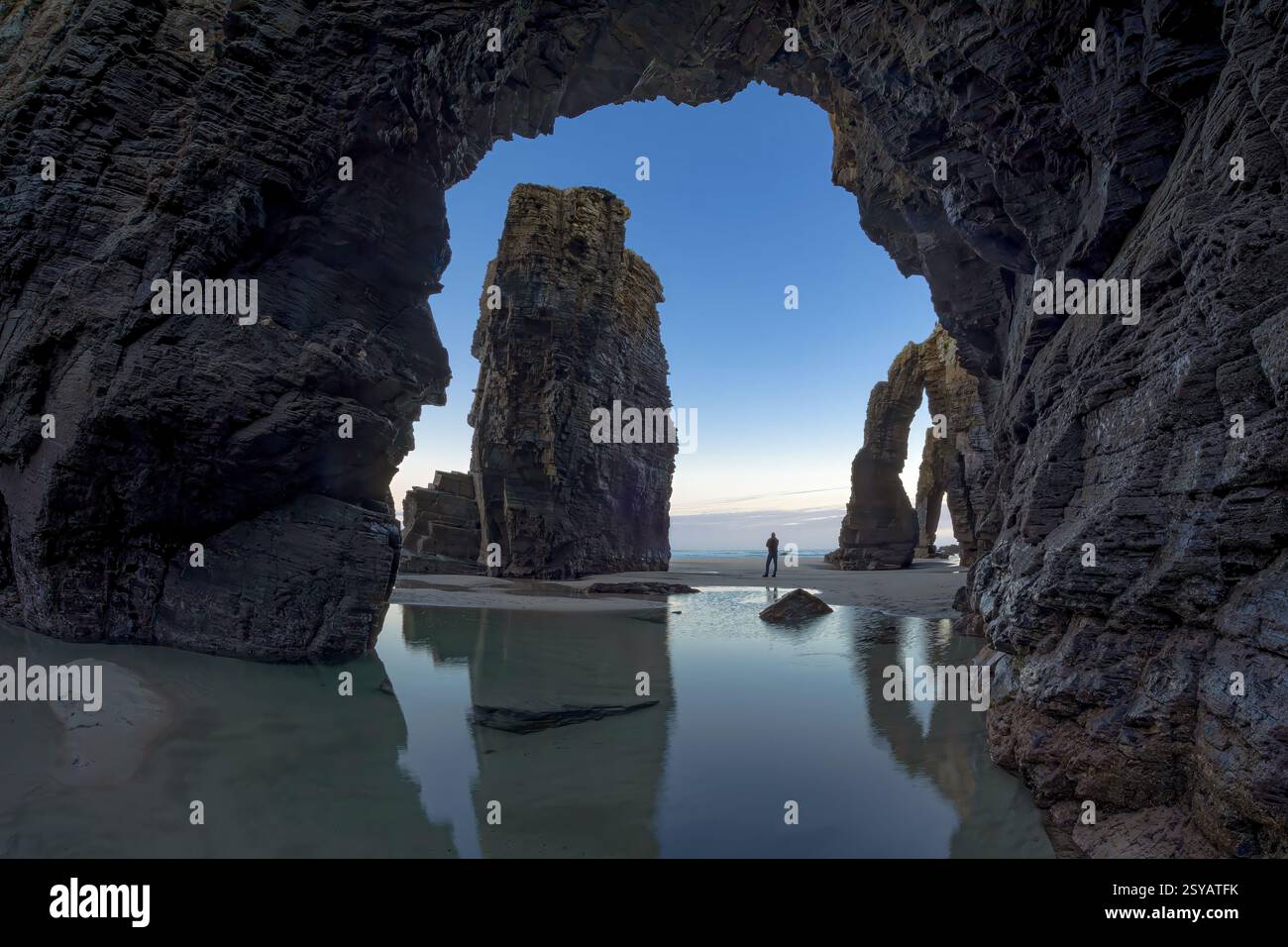 A person stands between towering sea arches, reflected in calm tidal ...