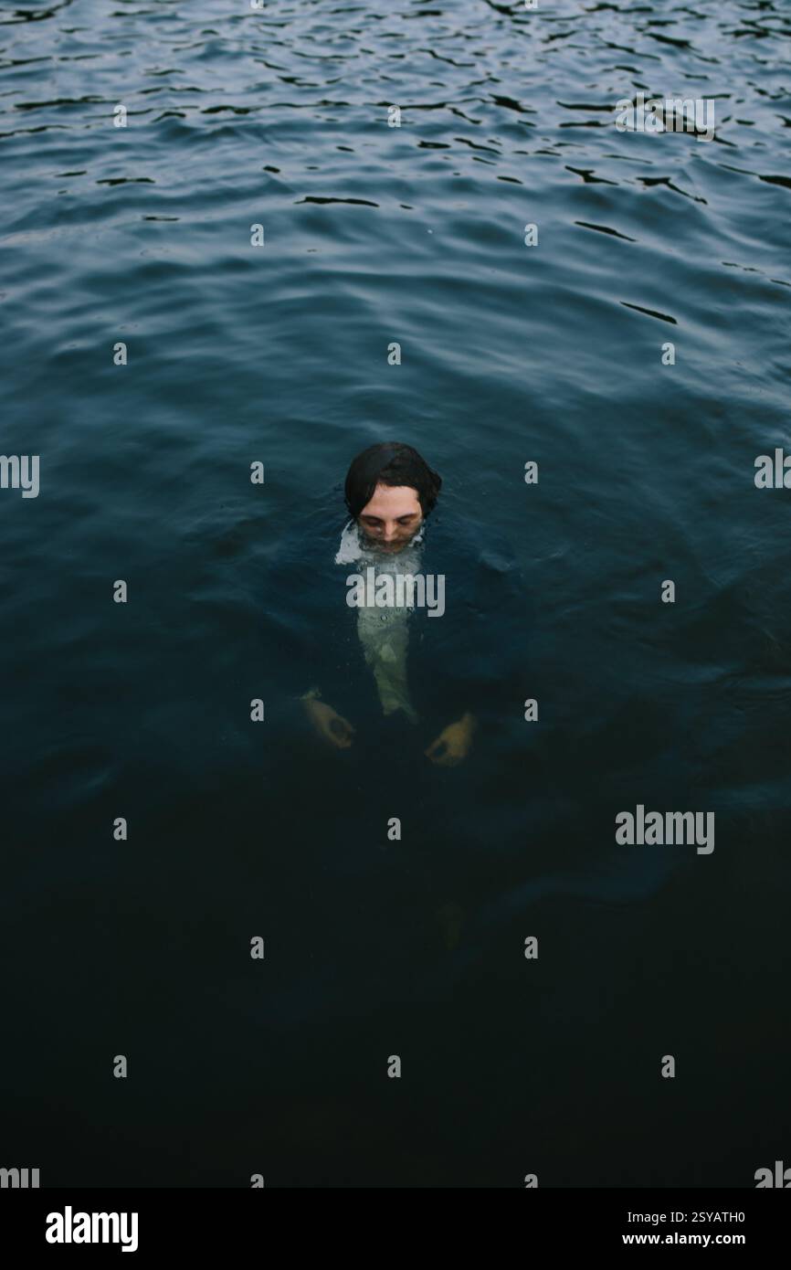 A man in formal clothing is partially submerged in deep, dark water. His expression and the ...