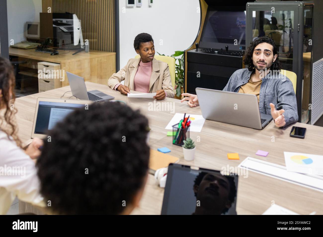 Diverse colleagues engage in a meeting, sharing ideas around a table with laptops and documents ...