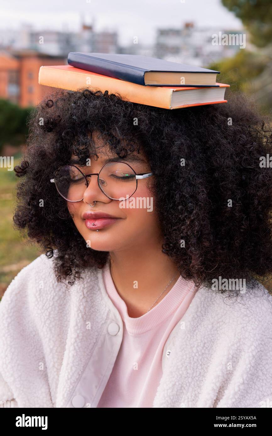 A young latin woman with curly hair and glasses balances two books on ...