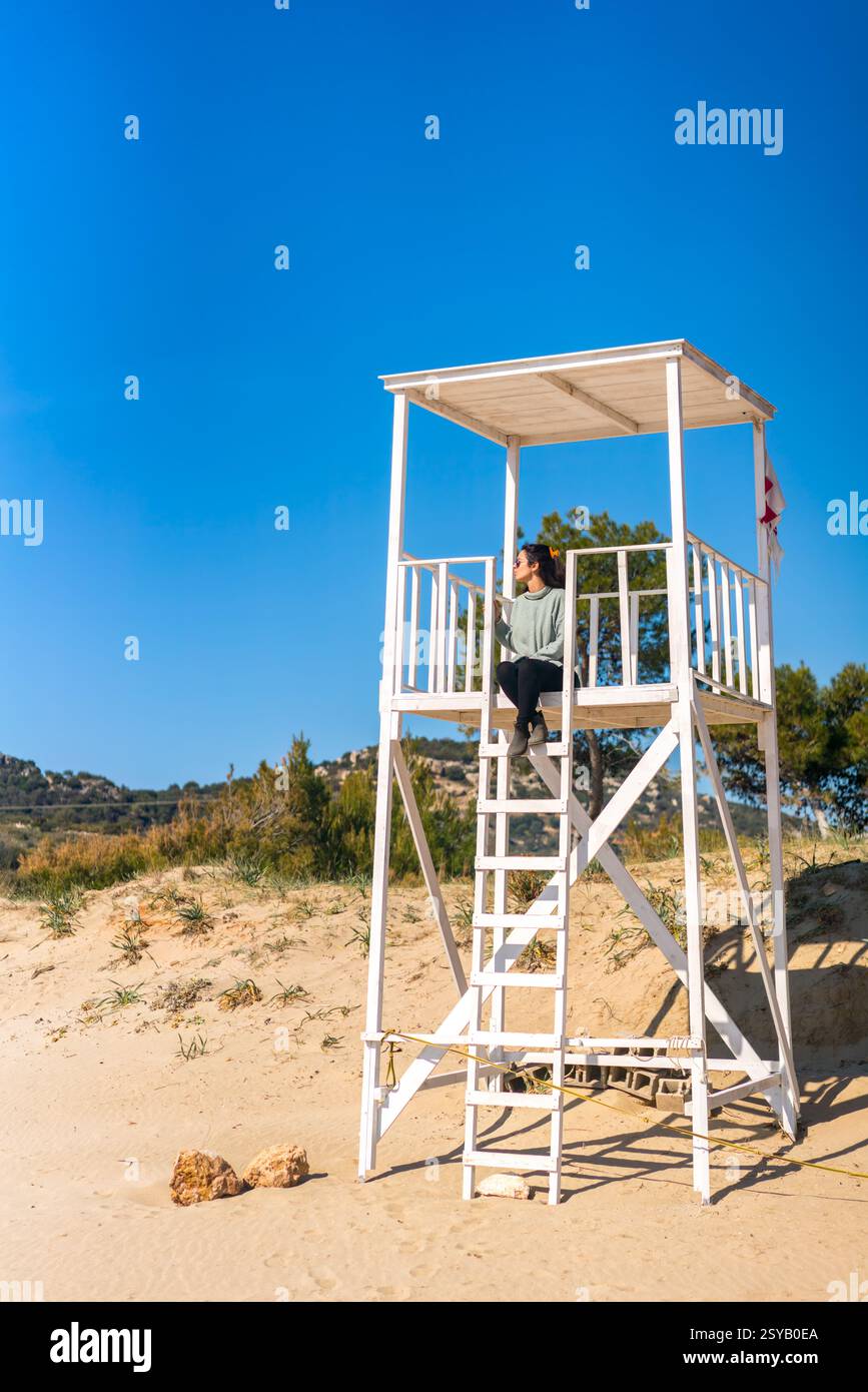 Woman sitting on a white lifeguard tower overlooking a sandy Greek ...
