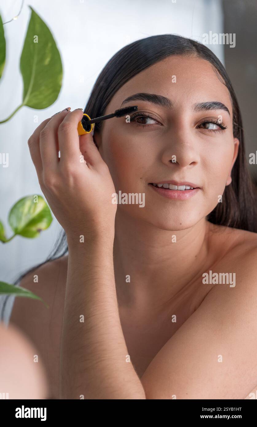 A woman smiling as she applies mascara in a brightly lit room. The ...