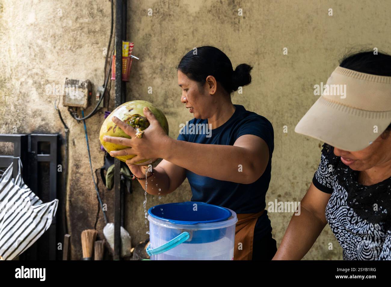 Two women skillfully open a coconut, letting its water drain into a ...