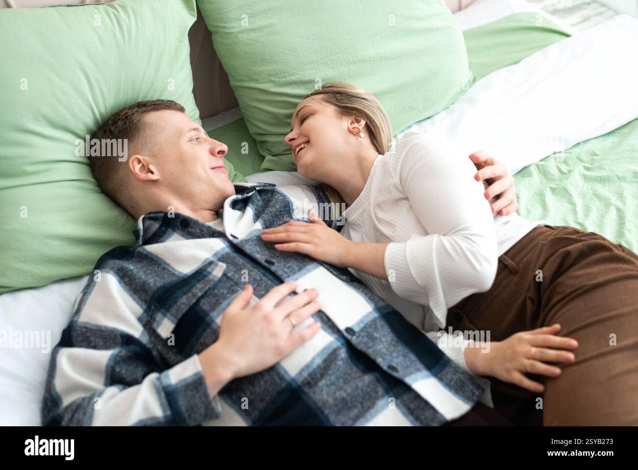 A young man and woman lie comfortably on a bed, sharing a close ...