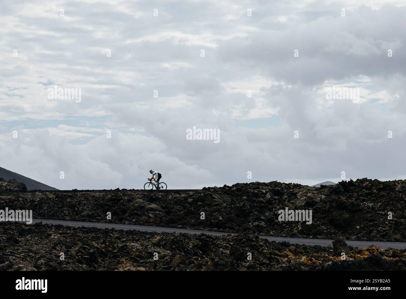 A lone cyclist riding on a volcanic road under a cloudy sky. The ...