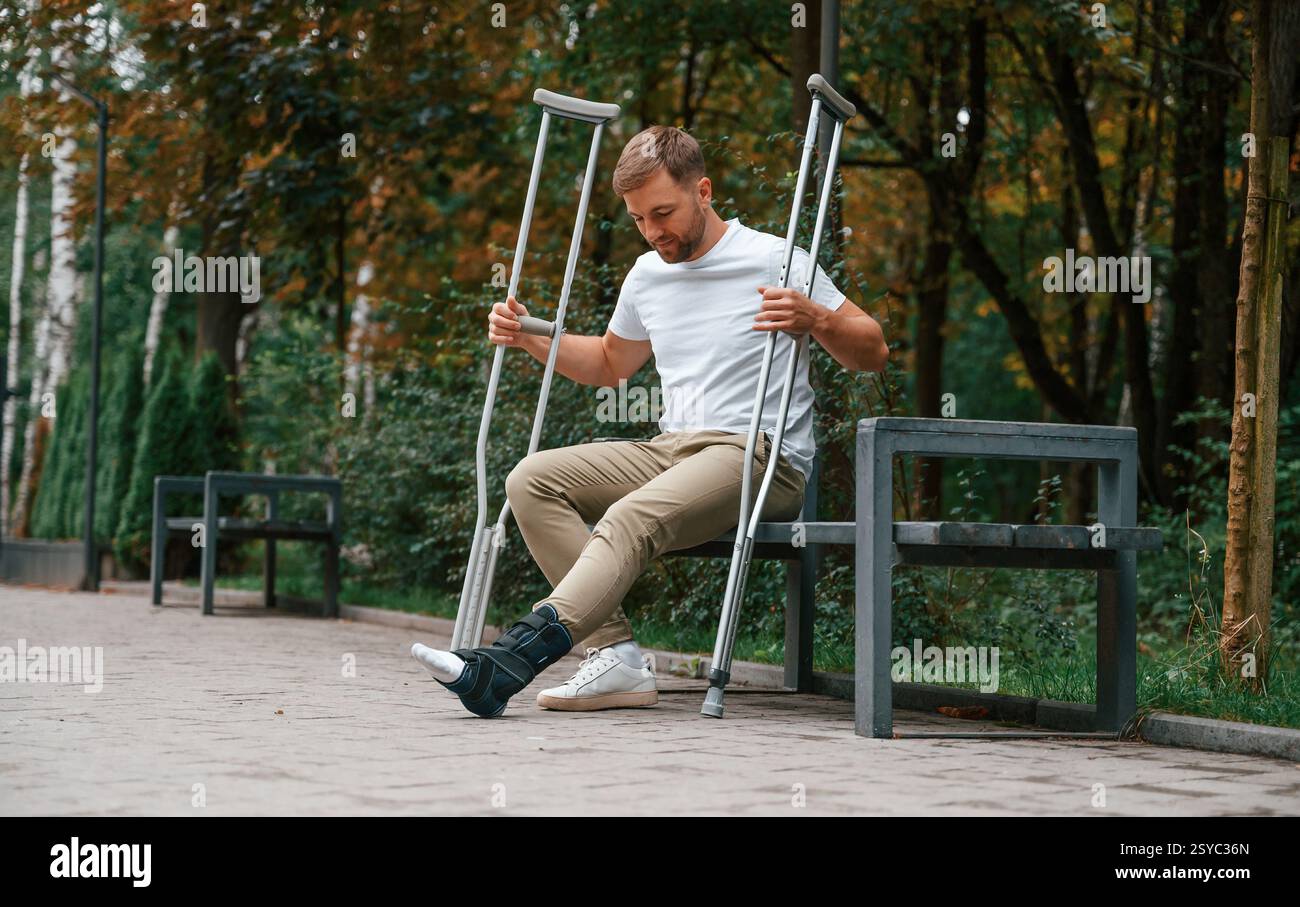Looking at the broken leg. Man with crutches is in the park outdoors ...
