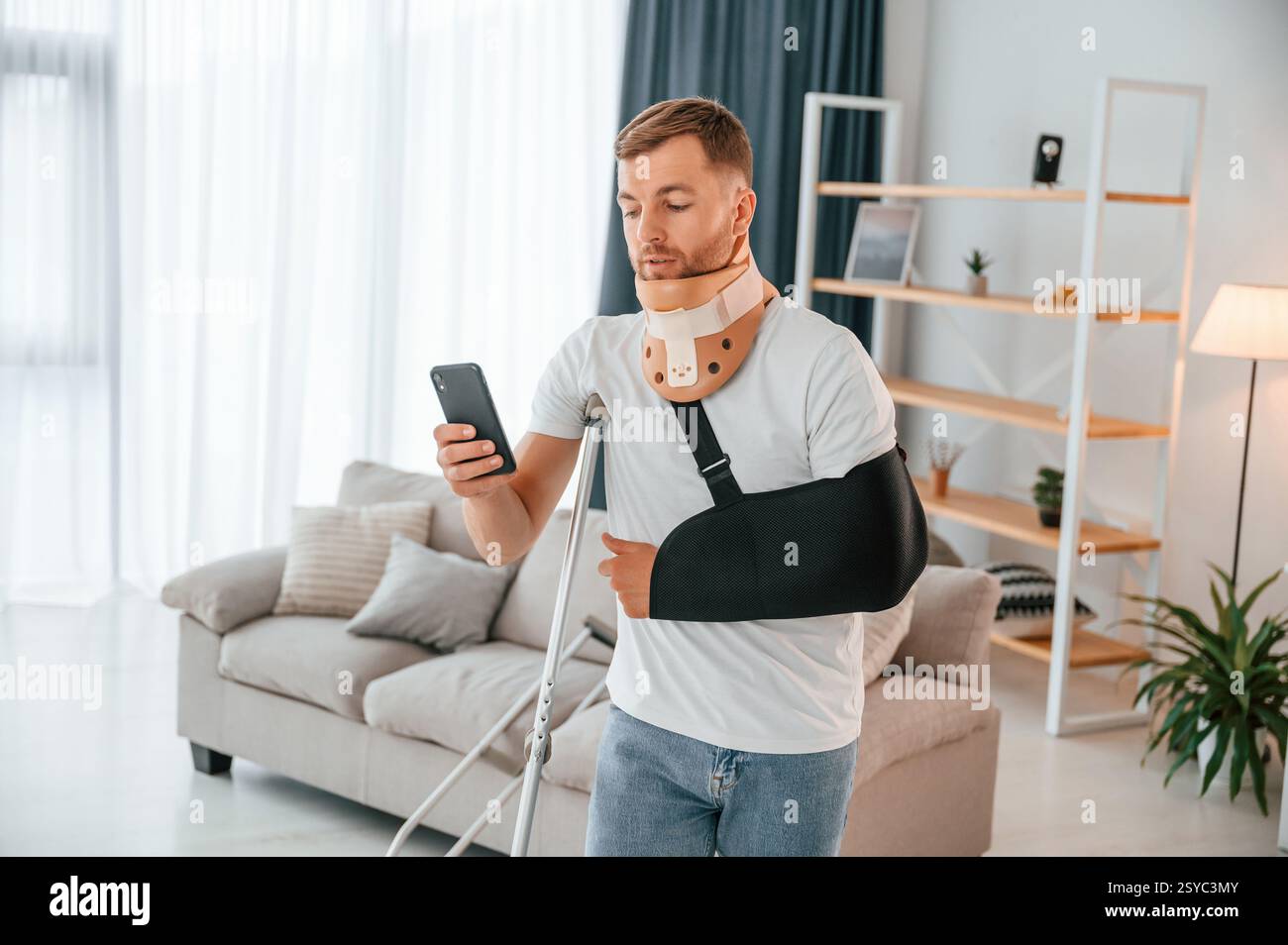 Using smartphone. Man with crutches is at home indoors. Having injuries ...