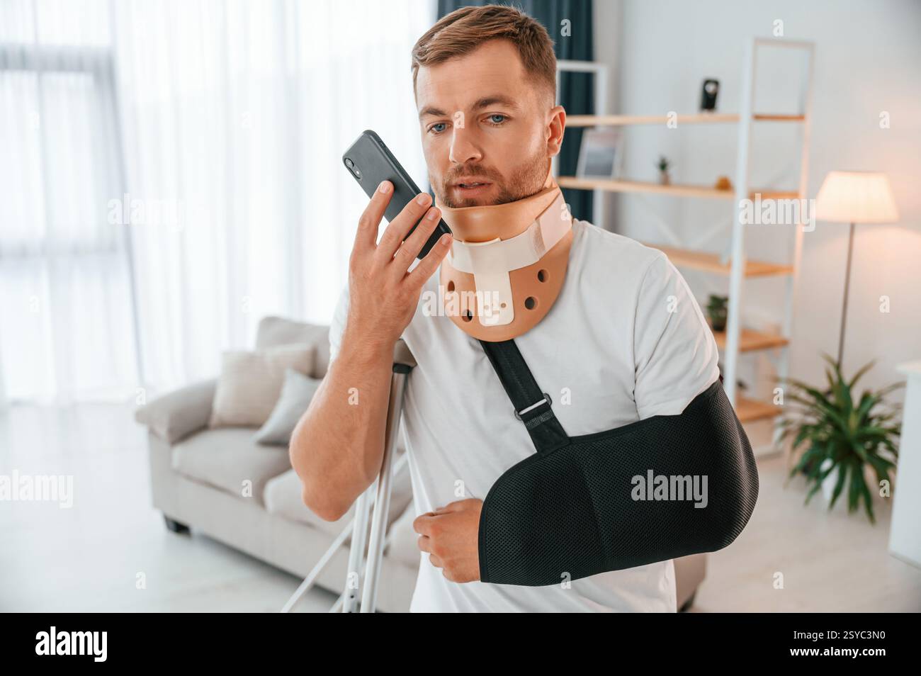Using smartphone. Man with crutches is at home indoors. Having injuries ...