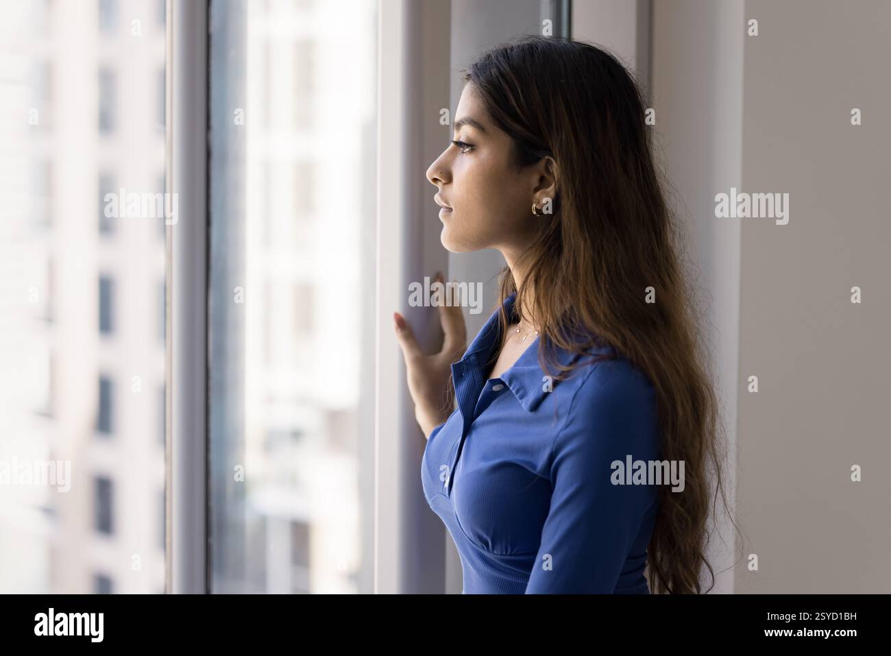 Indian woman standing by window, gazing outside with contemplative ...