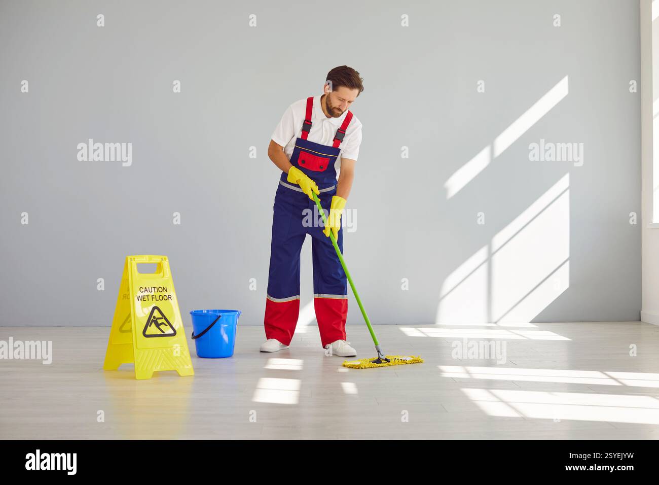 Male young janitor in uniform washing floor with caution wet floor sign ...