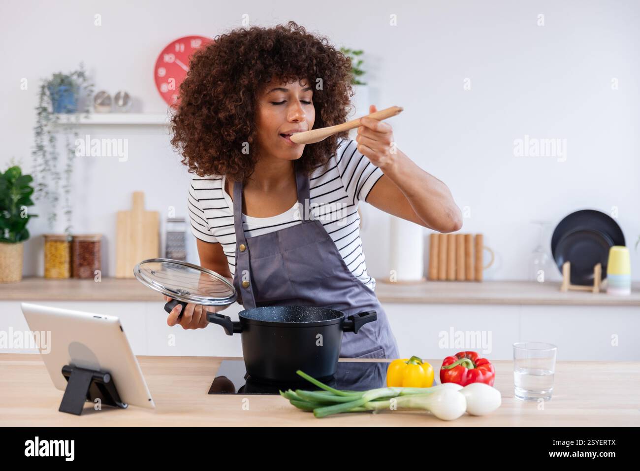 Young woman tasting food while following recipe on tablet in modern ...