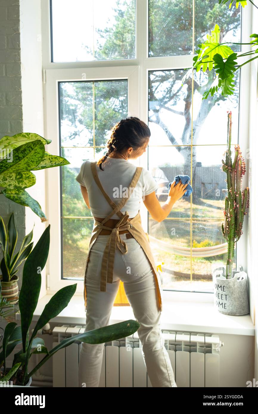 Woman in apron manually washes the window of house with rag cleaner and mop inside the interior ...