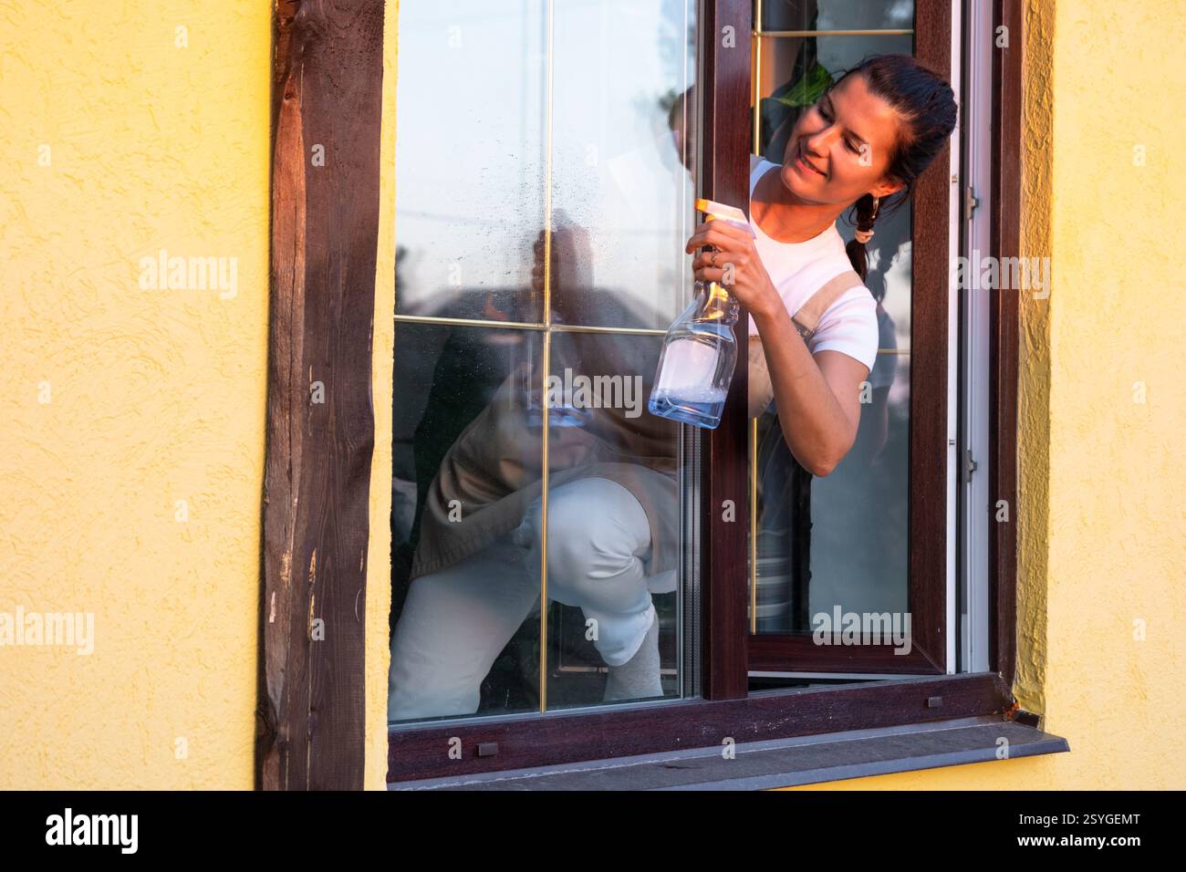 Woman in apron manually washes the window of the house with a rag with a spray cleaner and a mop ...