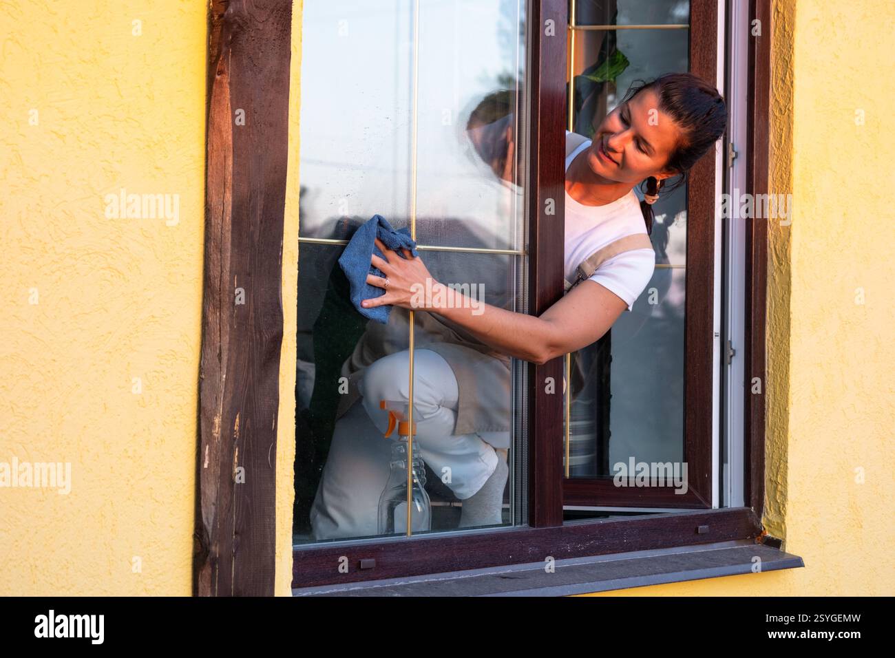 Woman in apron manually washes the window of the house with a rag with a spray cleaner and a mop ...