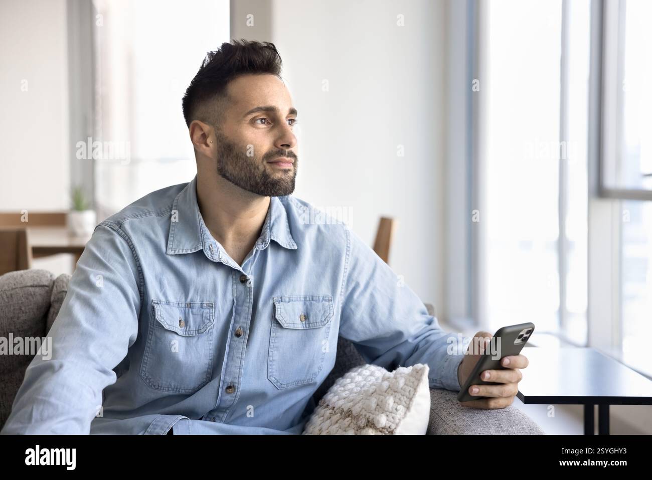 Man sit on couch, holds smartphone, gazing thoughtfully into distance ...