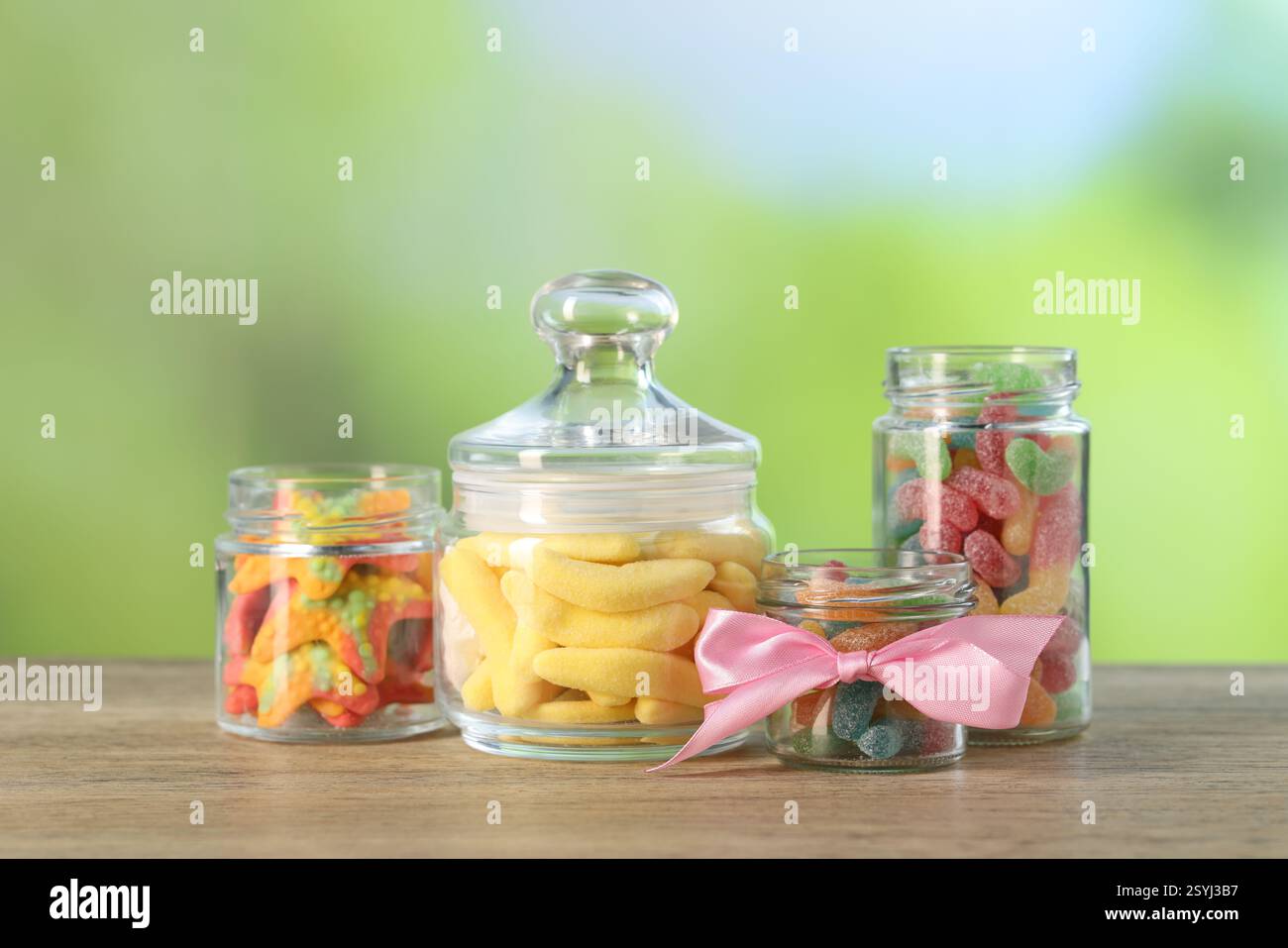 Tasty gummy candies in jars on wooden table against blurred green ...