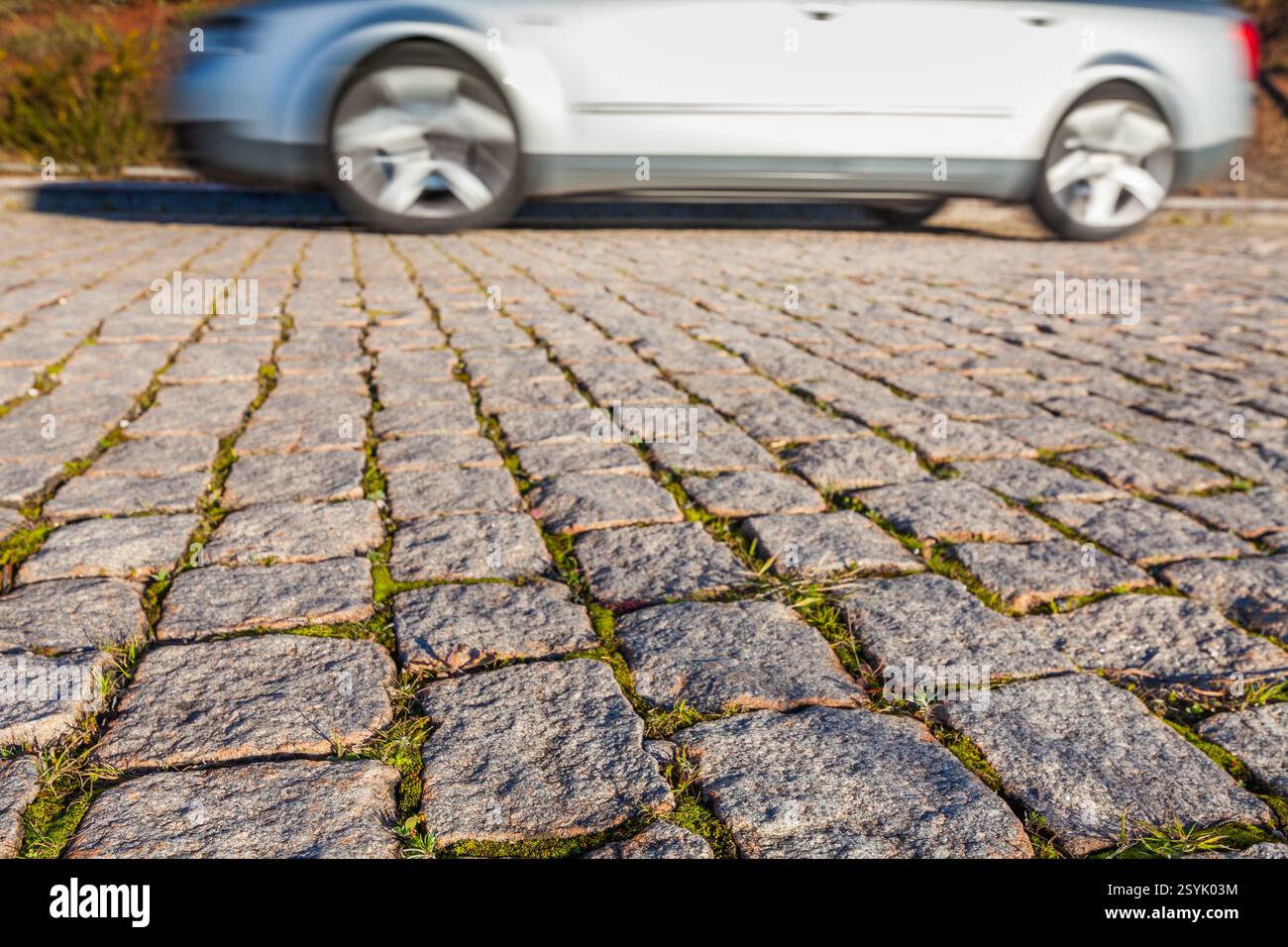 A blurred silver car drives over a textured cobblestone street, with a ...