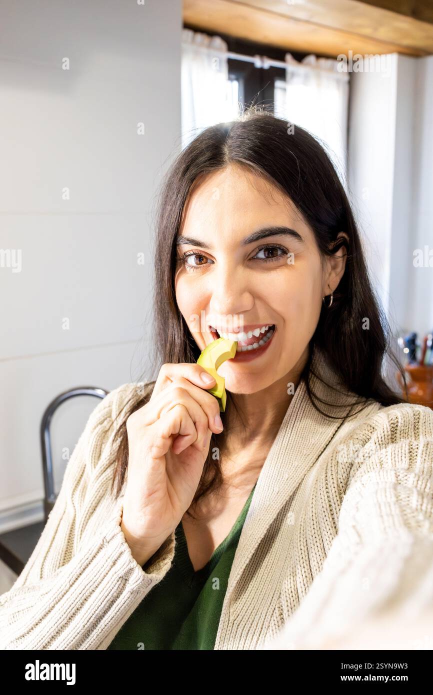 Happy woman taking a bite of fresh avocado in a bright kitchen. she enjoys a healthy snack while ...