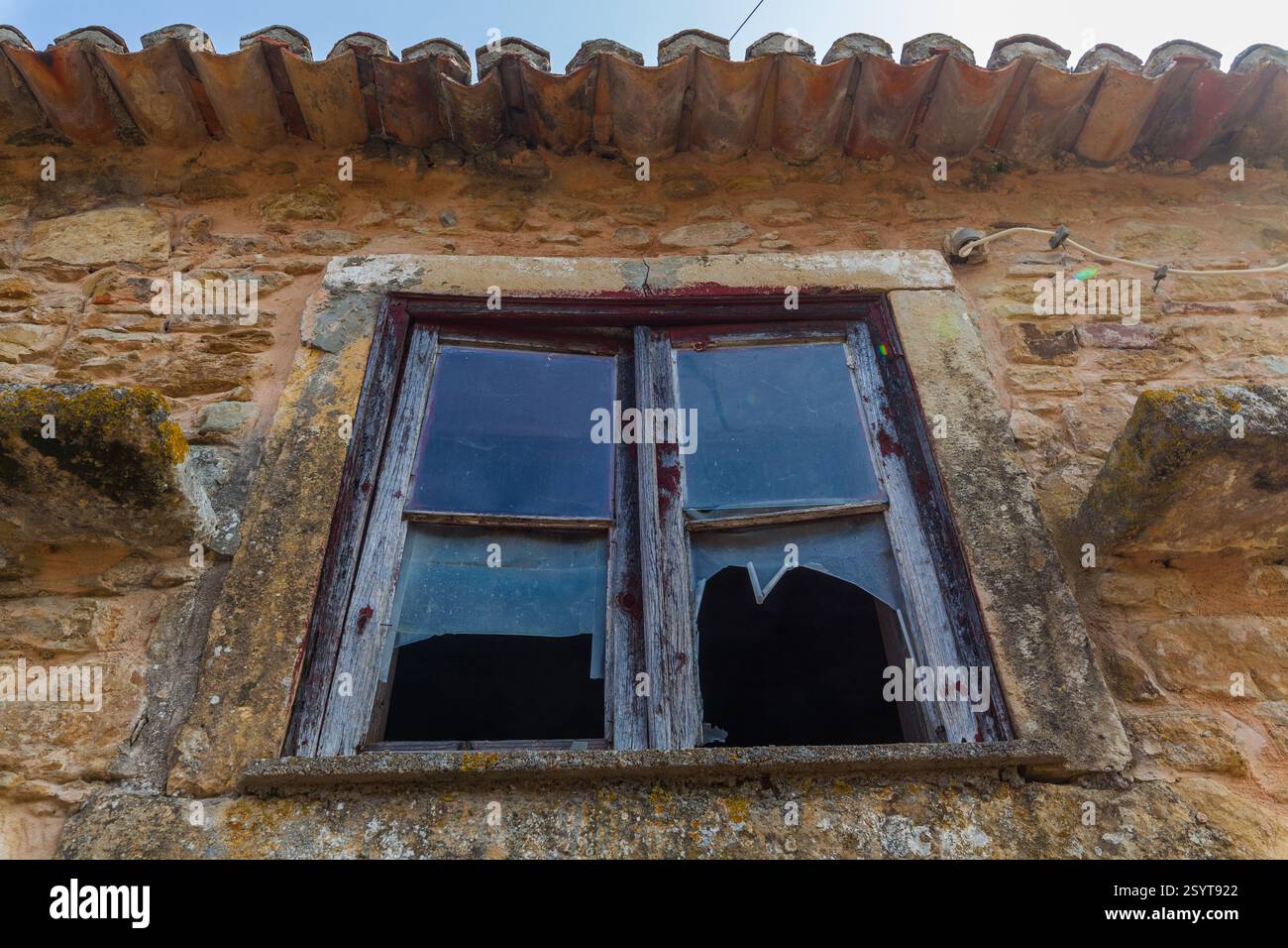 A weathered window with a broken pane is set in a rough stone wall ...