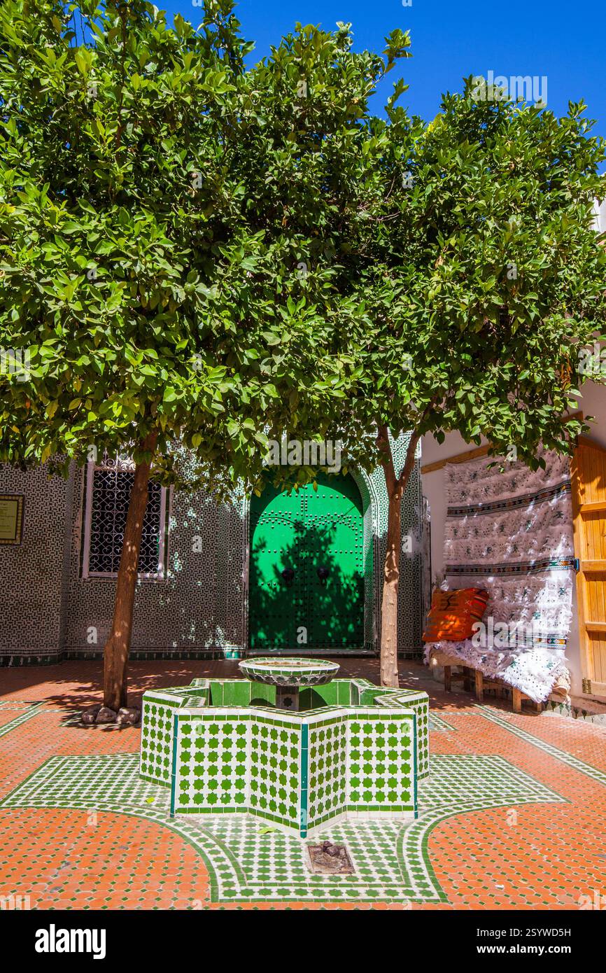 A serene courtyard featuring a tiled fountain, lush green trees, and a ...