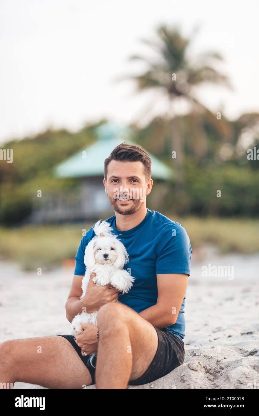 Young man with his cute white small dog maltese on the beach at sunset ...