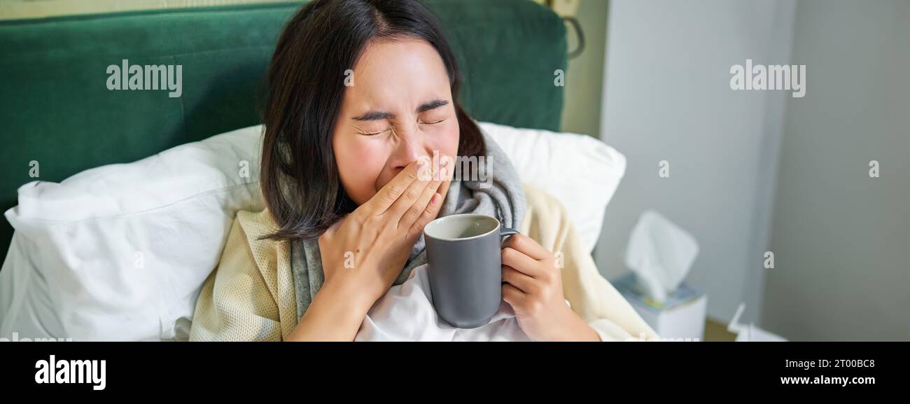 Close up of asian girl in bed, feeling sick, catching cold and sneezing ...