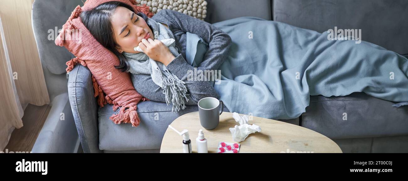 Sick sad korean woman lying on sofa, feeling unwell, catching cold, flu ...