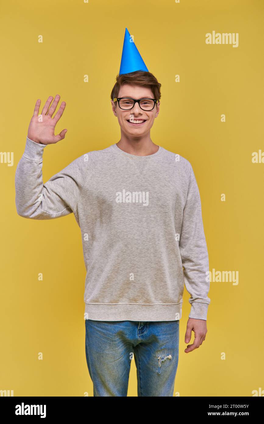 young smiley man in birthday hat waving his hand and smiling cheerfully ...