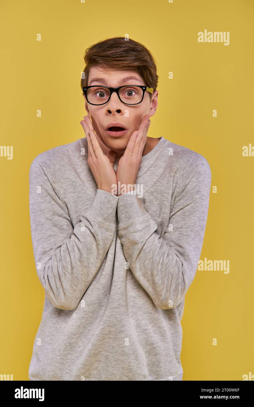 shocked young man in casual attire with glasses touching his cheeks ...