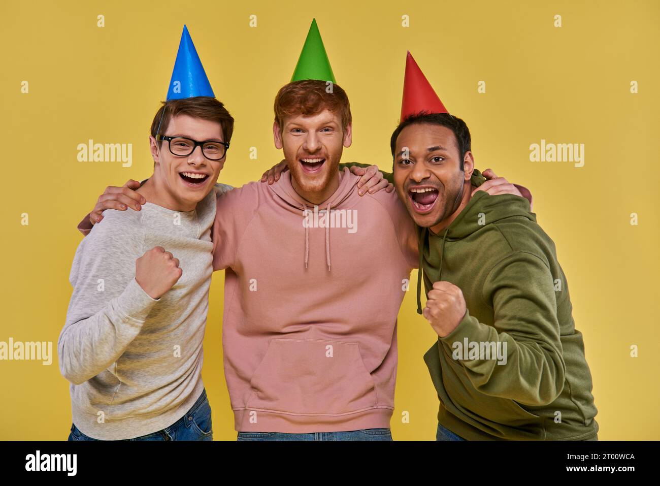 three multicultural cheerful friends in birthday hats hugging and ...