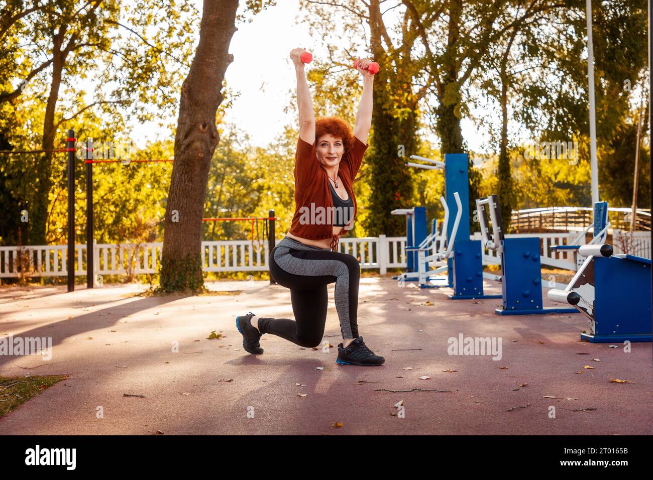 Adult Caucasian strength woman posing with dumbbells squatting in park ...