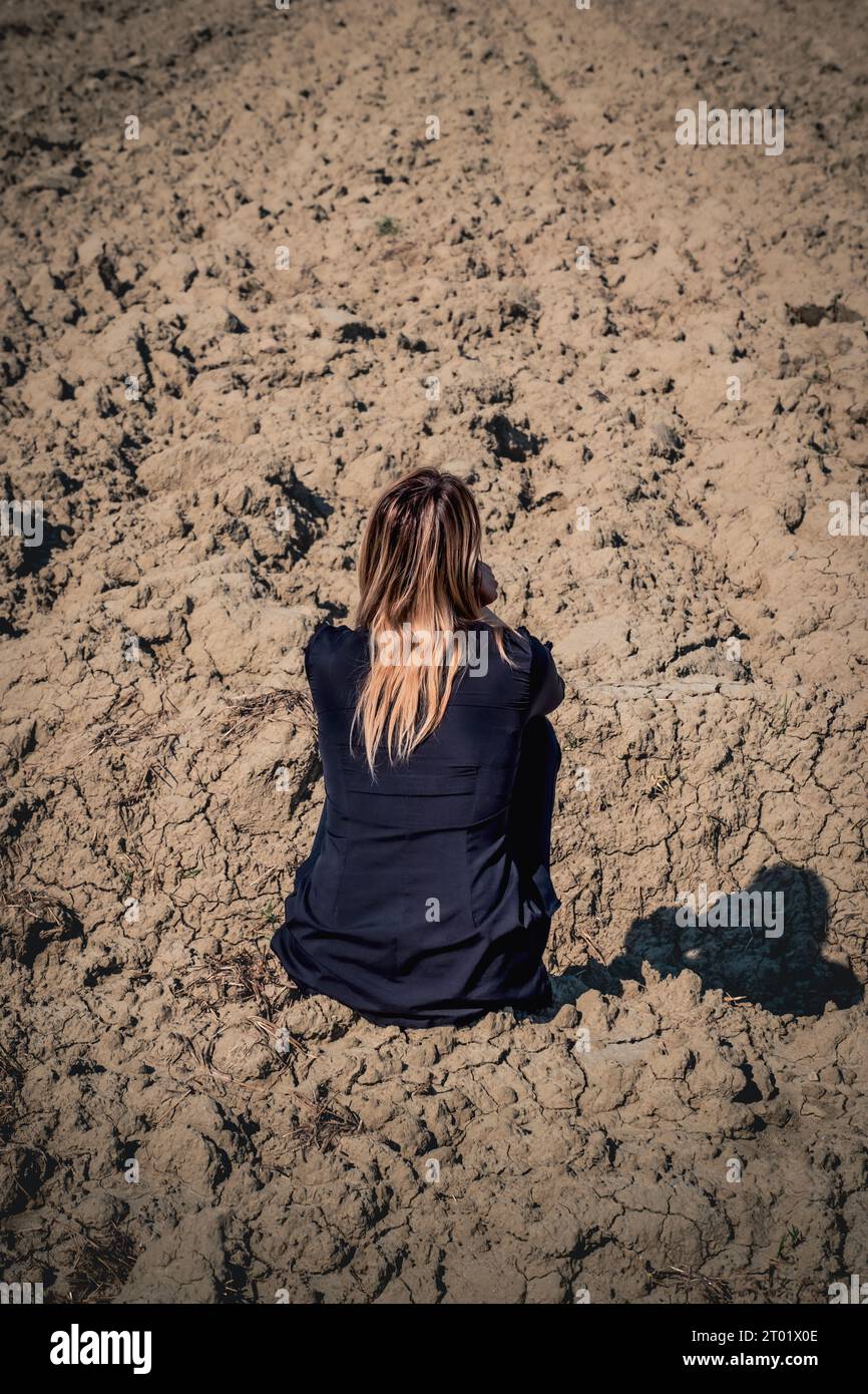 Thoughtful and sad woman sitting on the ground in a dry field due to ...