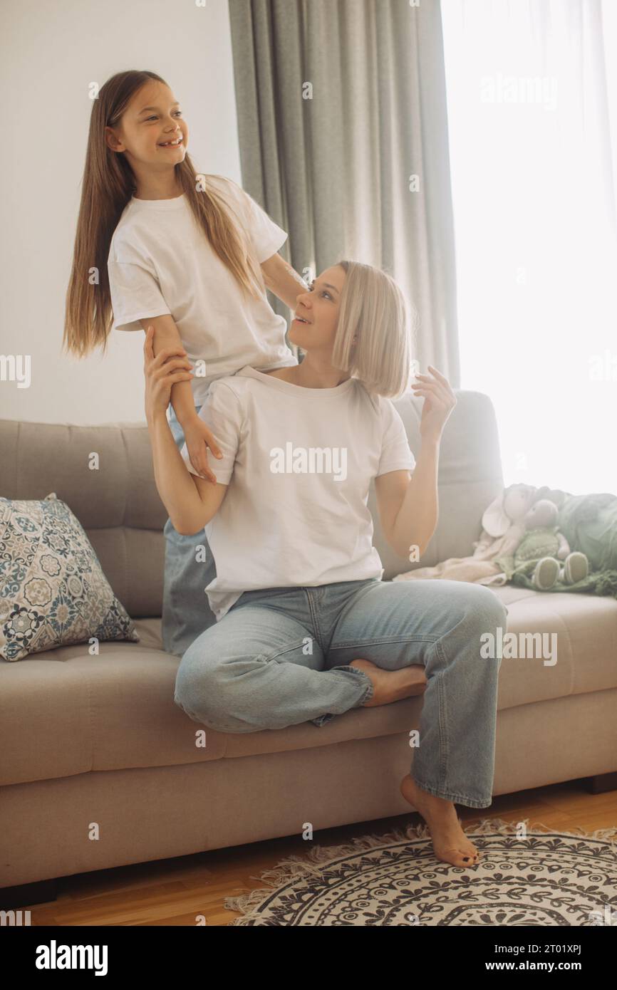 Cheerful mom playing laughing with little daughter on sofa, playful young mother having fun with ...