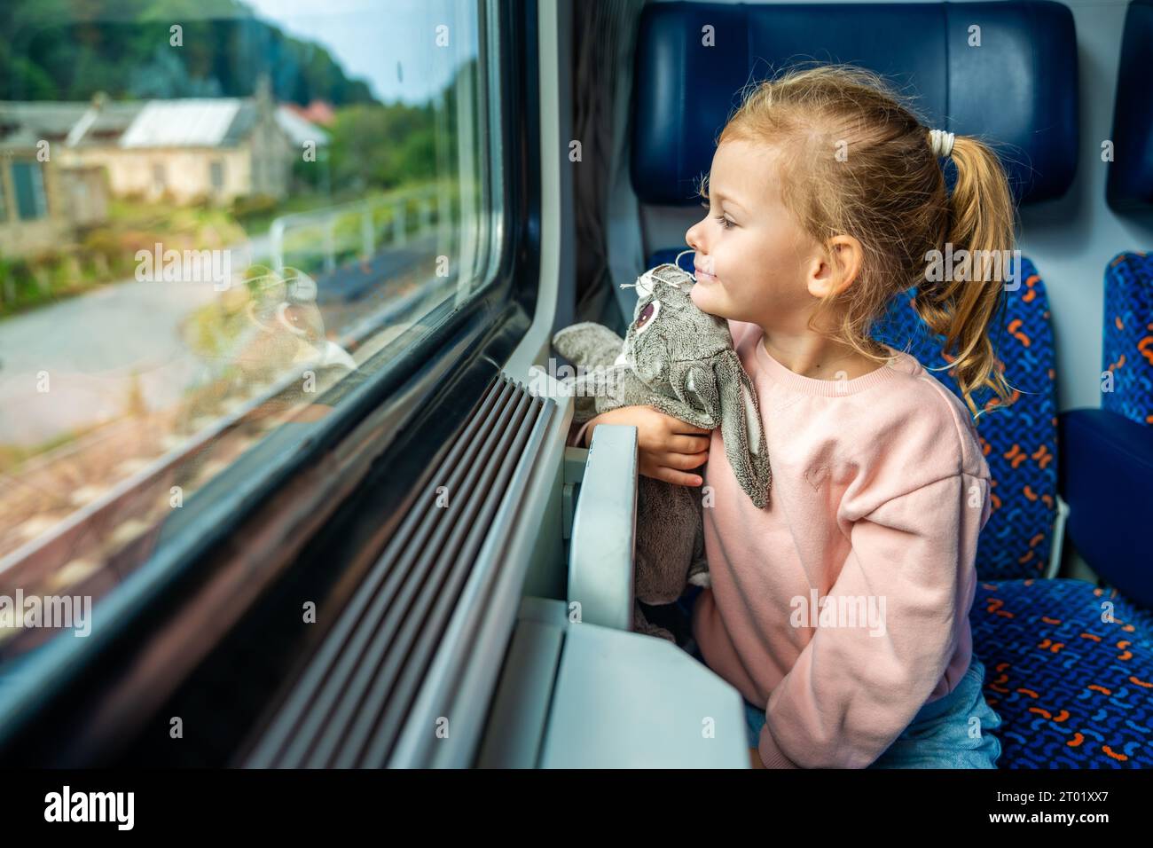 Smiling little girl with toy looking out train window outside, while it ...