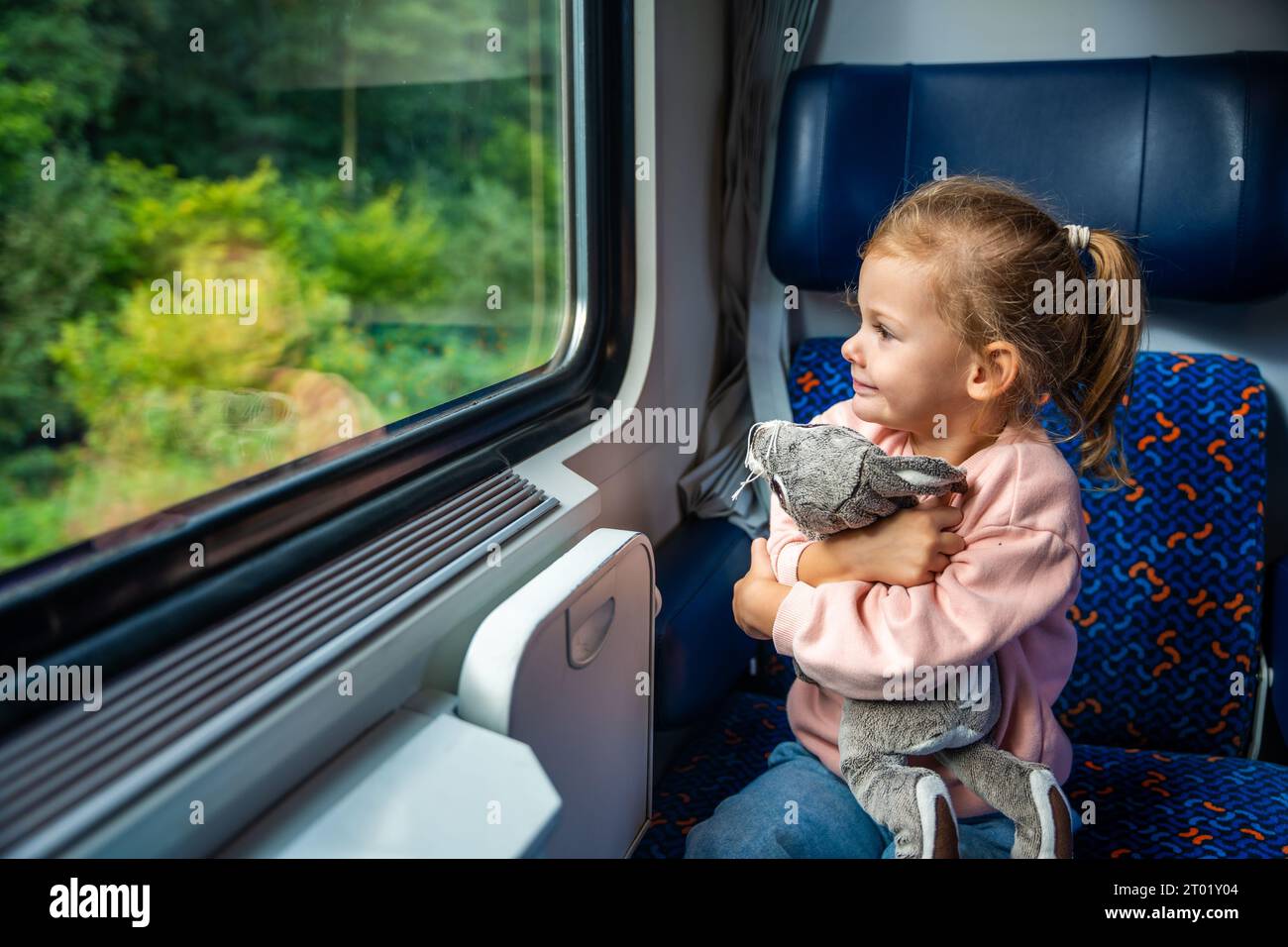 Beautiful little girl with toy looking out train window outside, while ...
