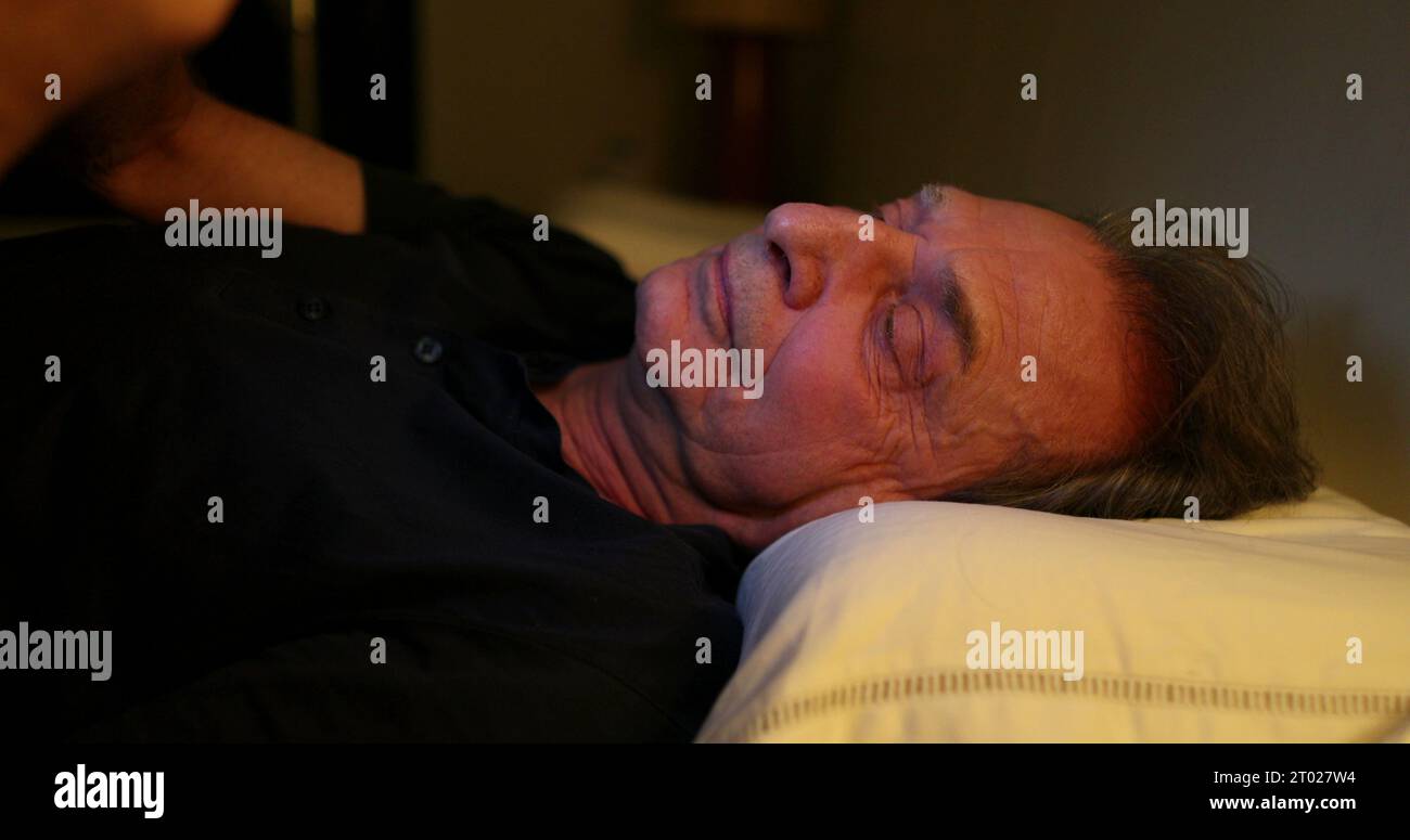 Older man picking up cellphone, laying down in bed looking at ...