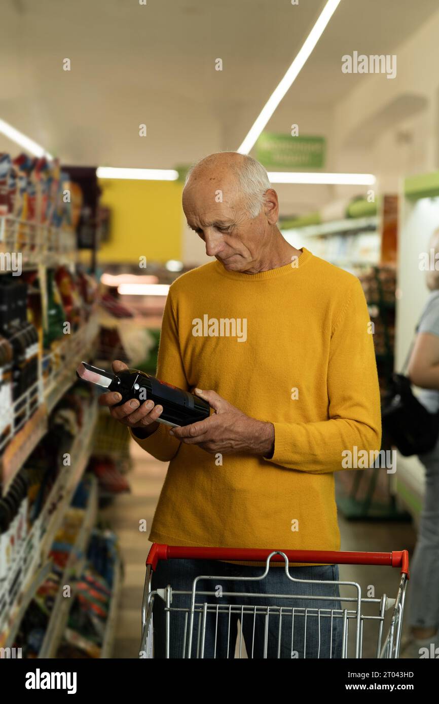 Old man chooses wine in supermarket. Elderly man reads labels on ...