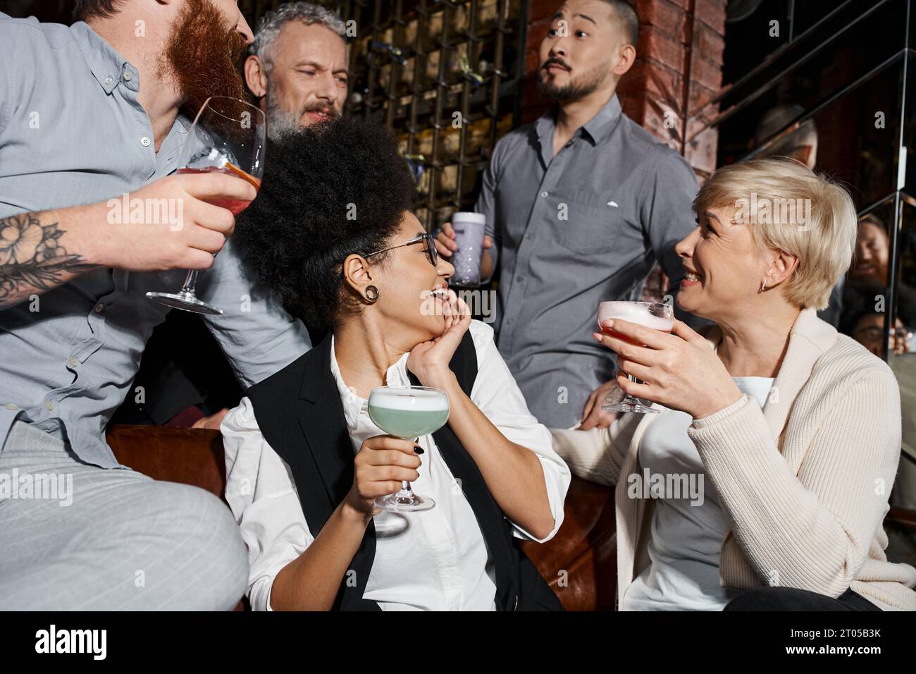joyful multicultural colleagues drinking cocktails and talking in bar ...