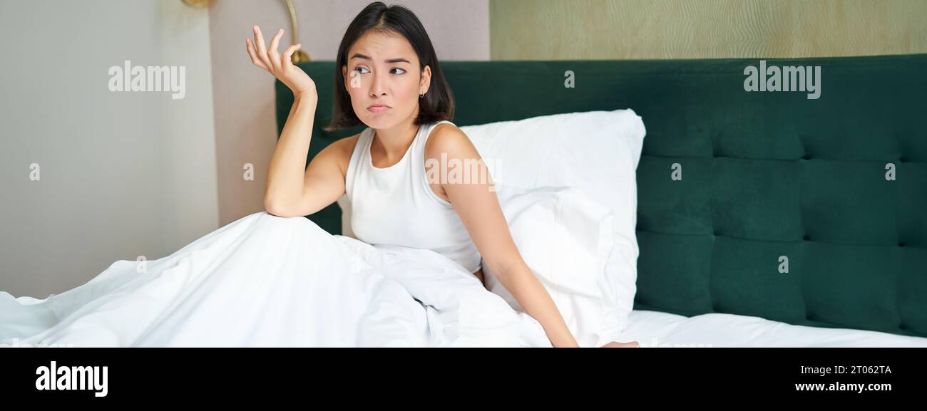 Portrait of puzzled asian woman lying in bed covered with warm duvet ...