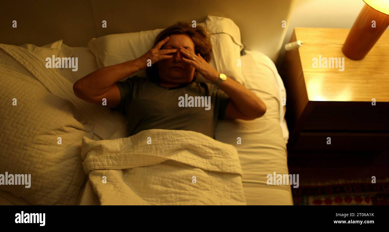 Sleepless senior woman turning light on and rubbing face with hands ...