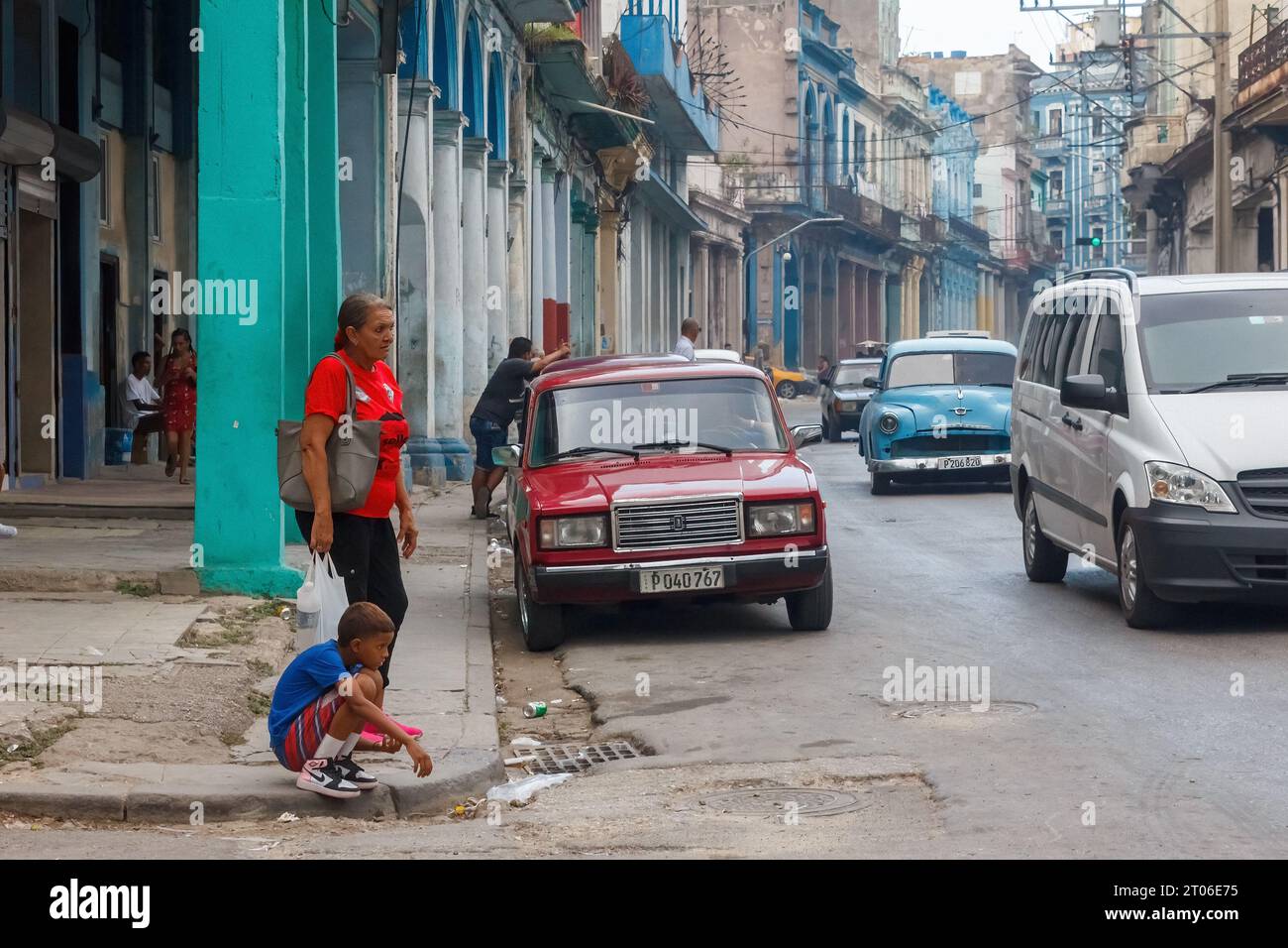 A Cuban woman and a child stand on a broken city sidewalk by an avenue ...