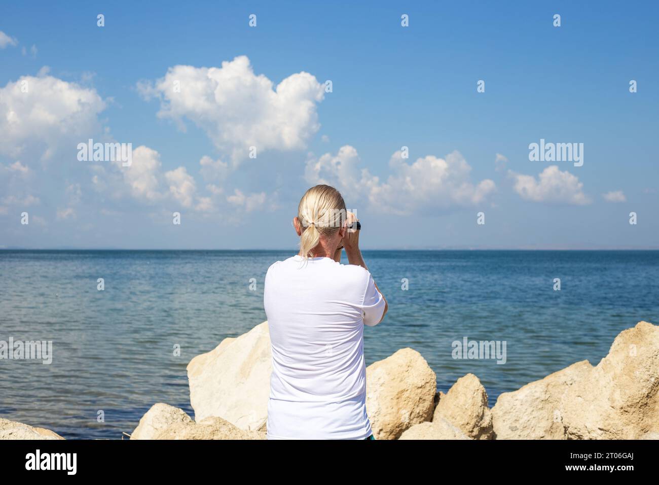A blond man with hair in a ponytail films a seascape with a video ...