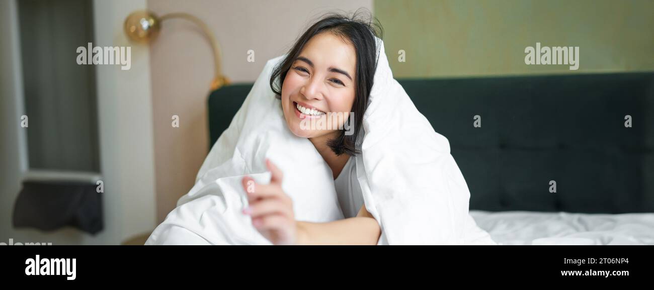 Beautiful asian woman sitting on bed, covered with white duvet, smiling ...