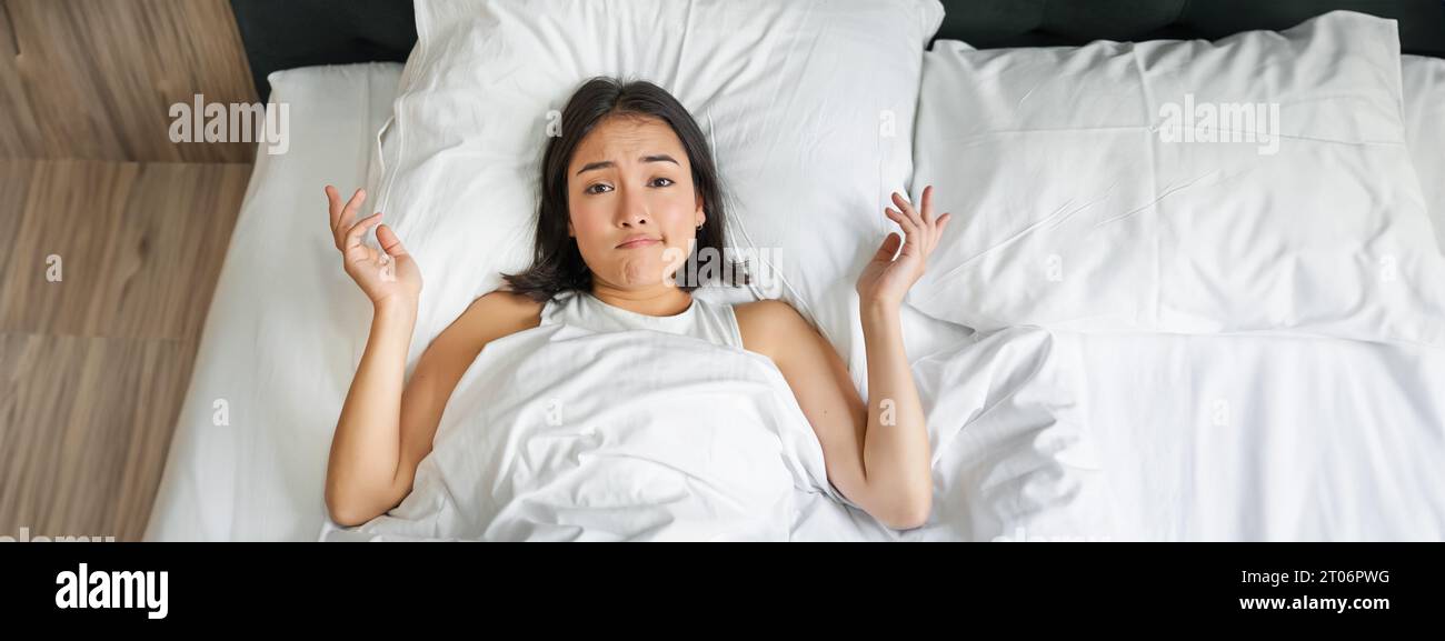 Portrait of asian girl lying in her bed and shrugs, looks puzzled ...