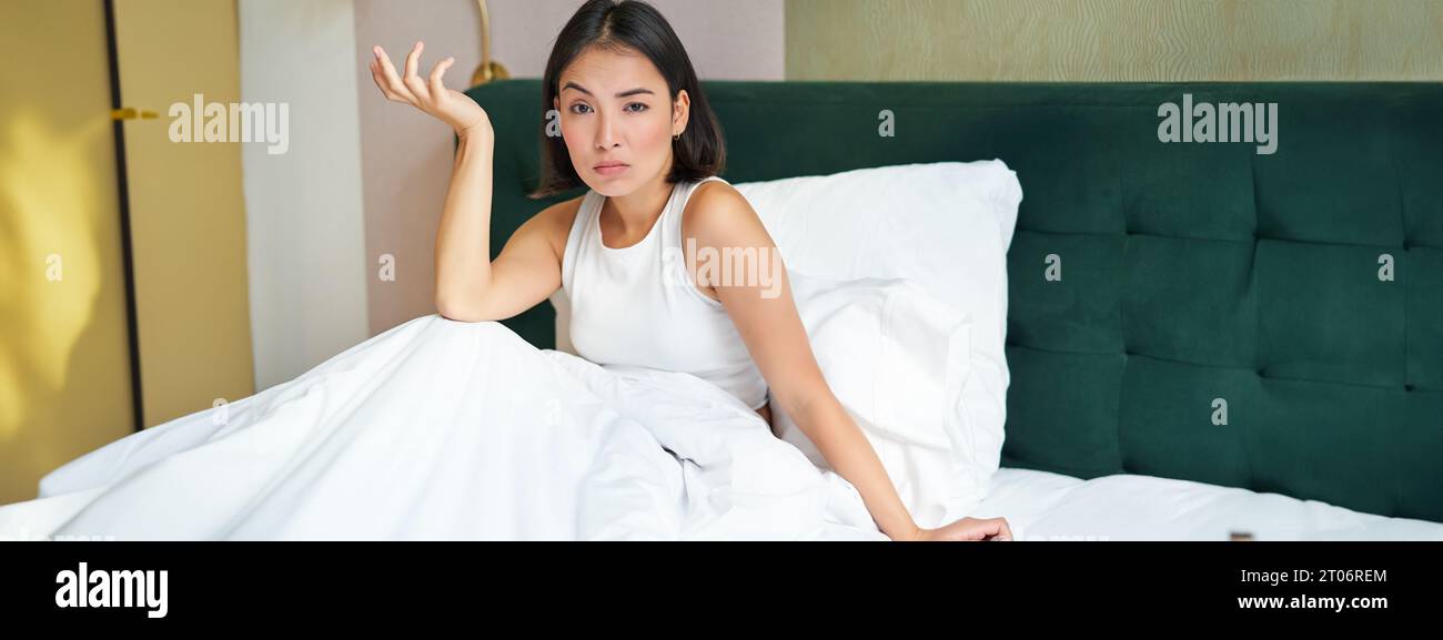 Portrait of puzzled asian woman lying in bed covered with warm duvet ...