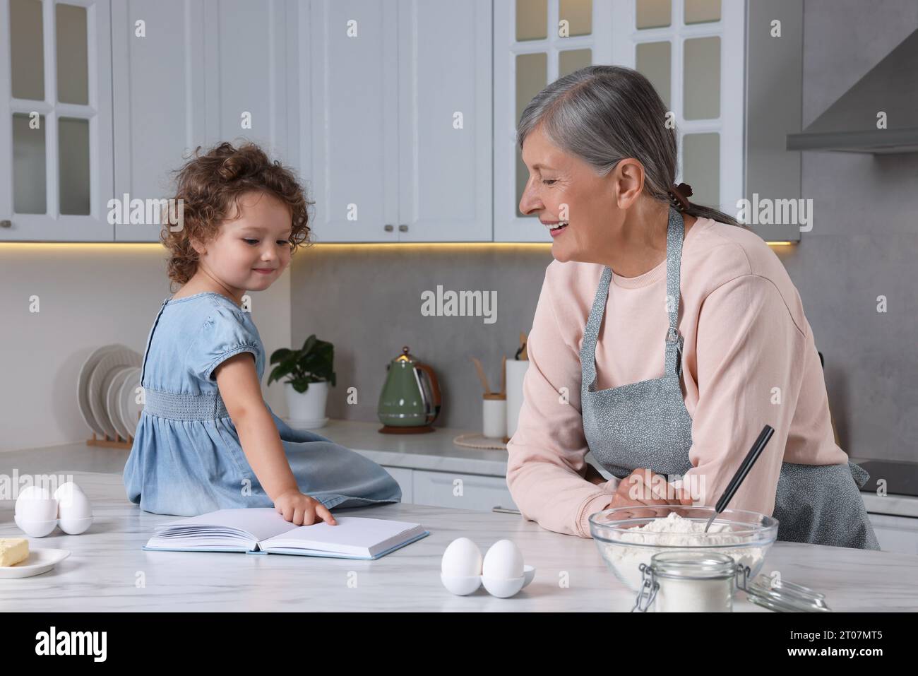 Cute little girl with her granny cooking by recipe book in kitchen ...