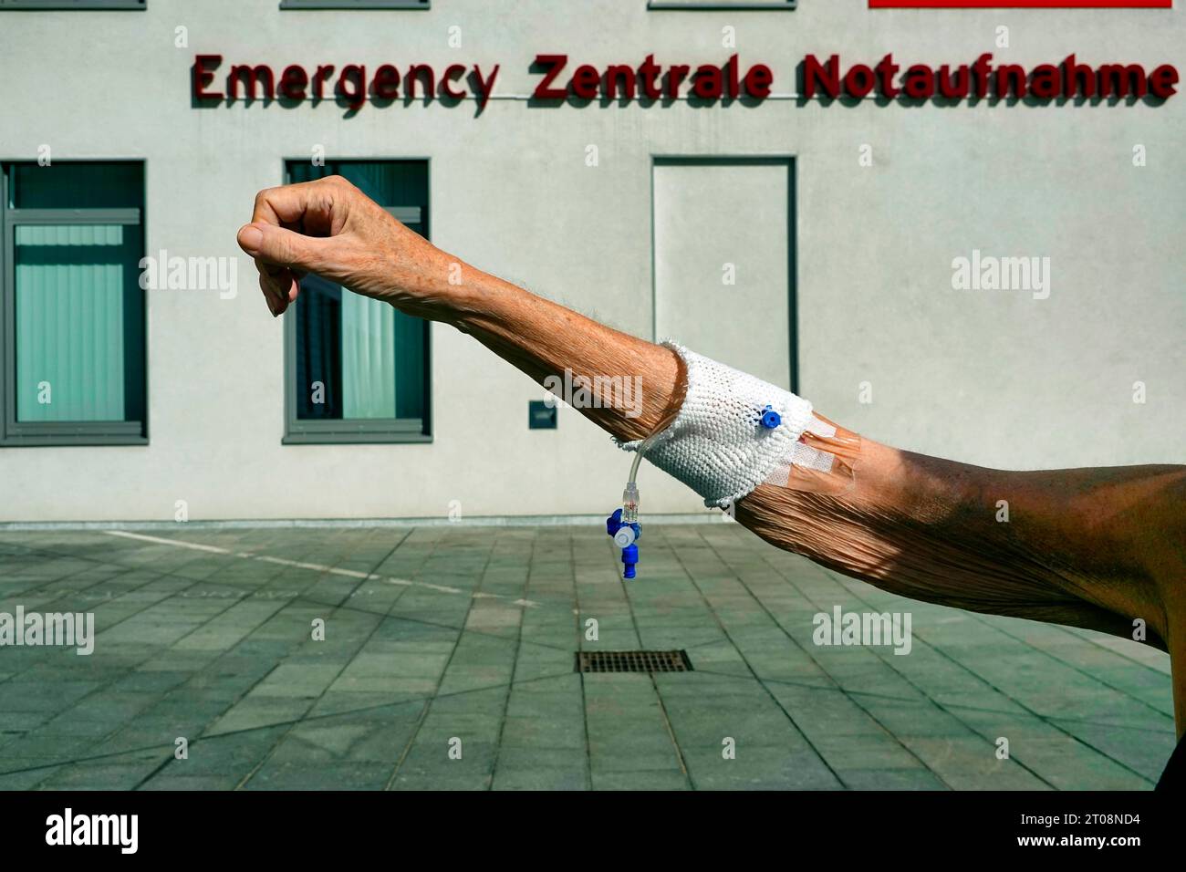 A raised emaciated hand with a bandage and a brown line in place, the ...