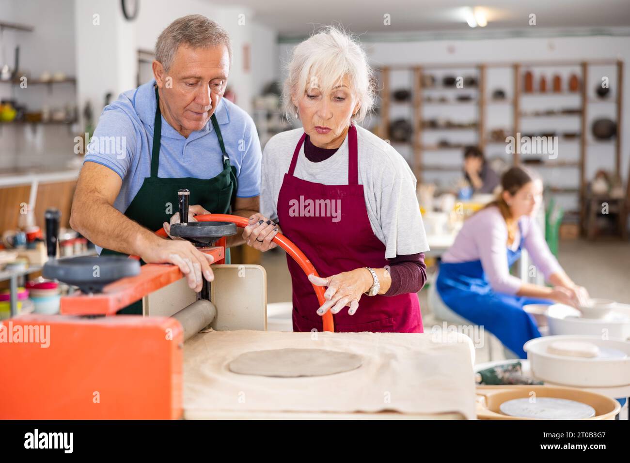 Elderly potter teaches woman how to roll clay on a craft machine clay ...